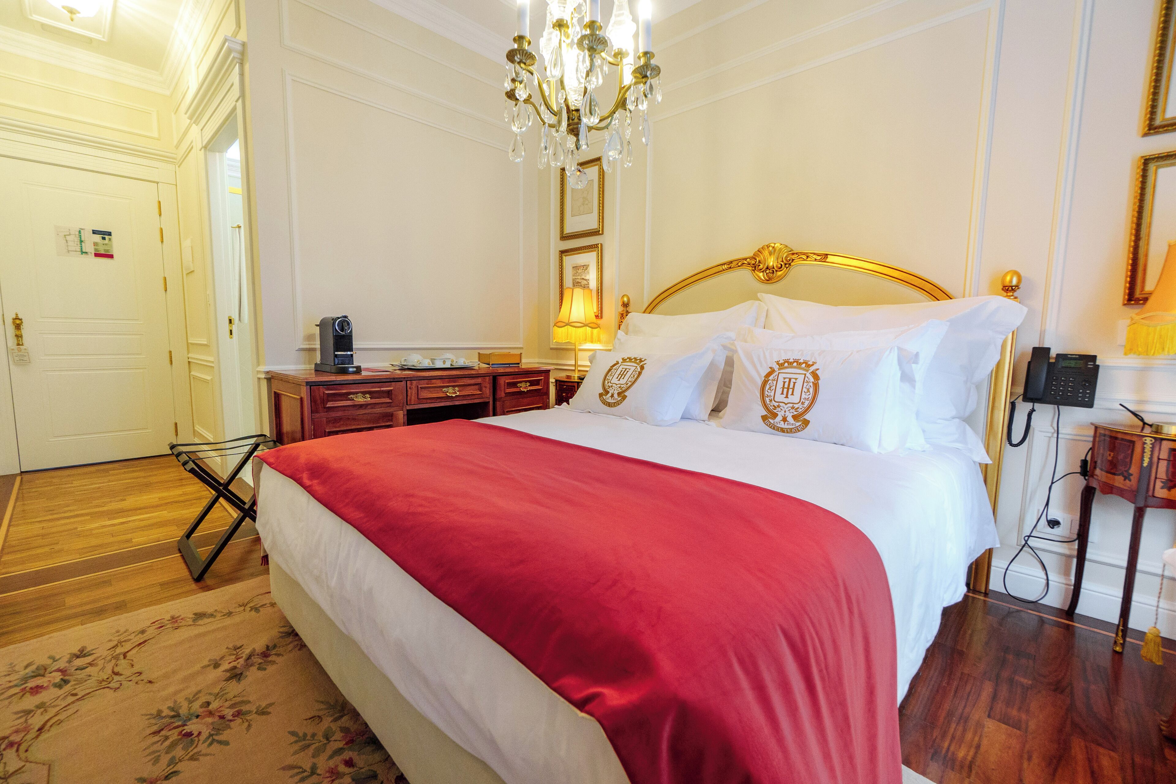 Quarto de hotel com uma cama grande, um candelabro, uma escrivaninha com telefone e uma porta.
