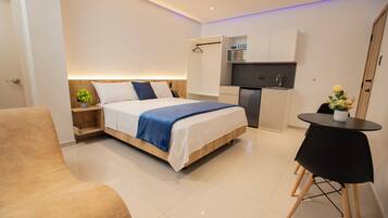 Quarto de hotel moderno com cama, mesa de cabeceira, pequena escrivaninha e cozinha compacta.