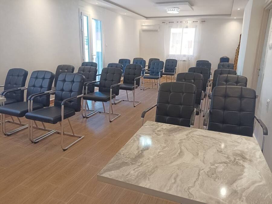 Uma sala de conferências com fileiras de cadeiras pretas, uma mesa de mármore e grandes janelas com cortinas.