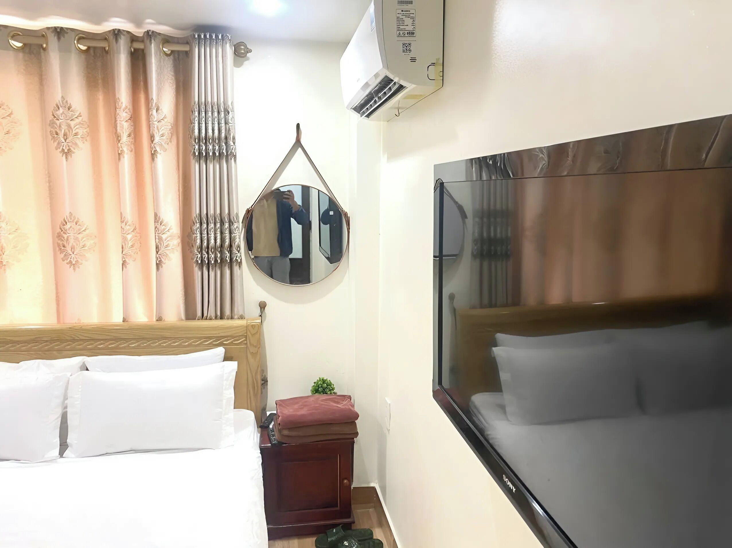 Quarto de hotel com uma cama, mesa de cabeceira, espelho e uma grande tela de televisão.