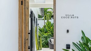 Uma entrada moderna com porta de madeira, um cachepot de concreto com uma planta grande e uma parede com o texto "VILLA COCO.NUTS".