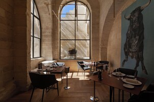 Uma área de refeições com mesas e cadeiras, uma grande janela em arco e um mural de um touro na parede.