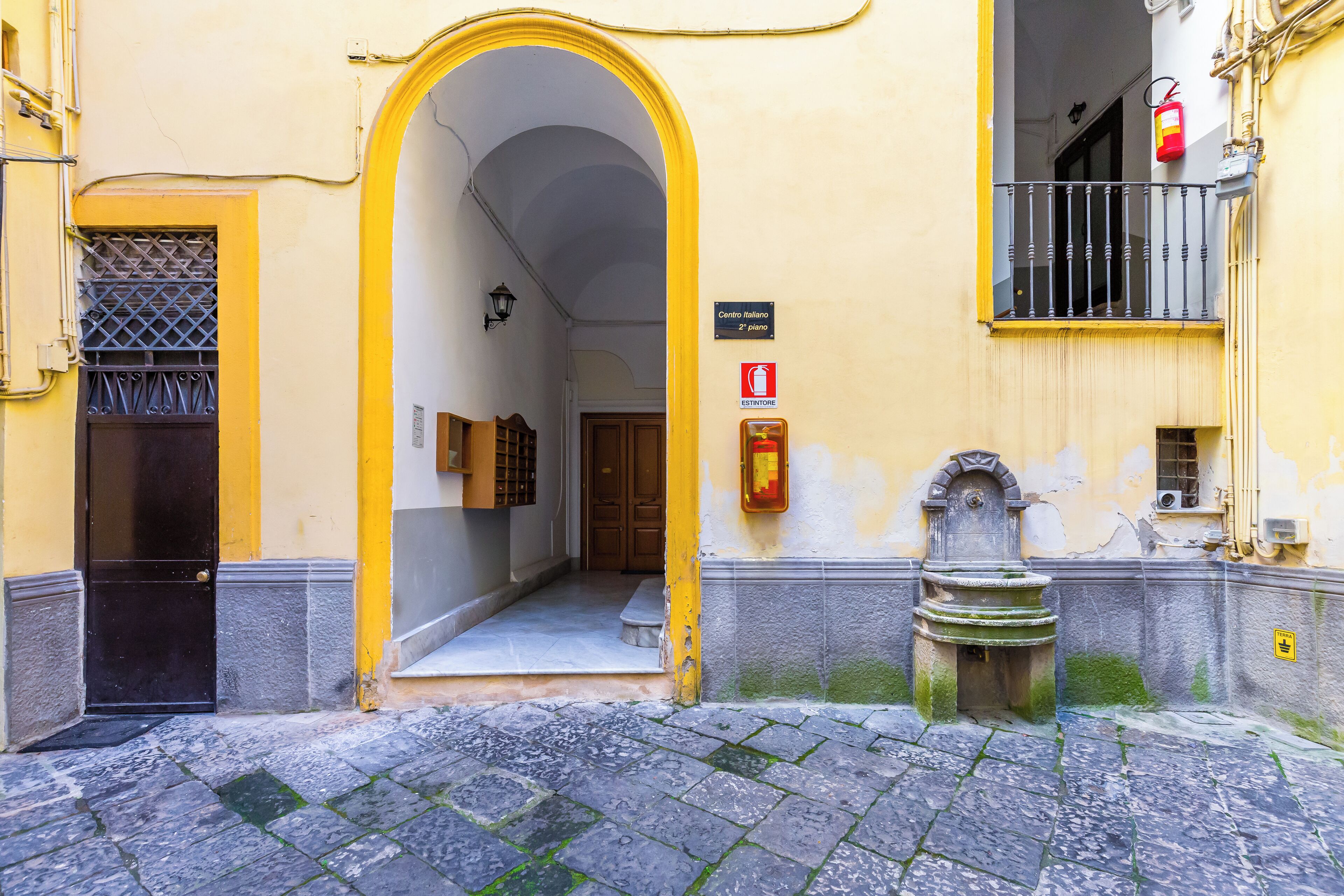 Uma rua estreita com um edifício amarelo, uma porta preta e uma fonte de pedra.