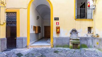 Uma rua estreita com um edifício amarelo, uma porta preta e uma fonte de pedra.