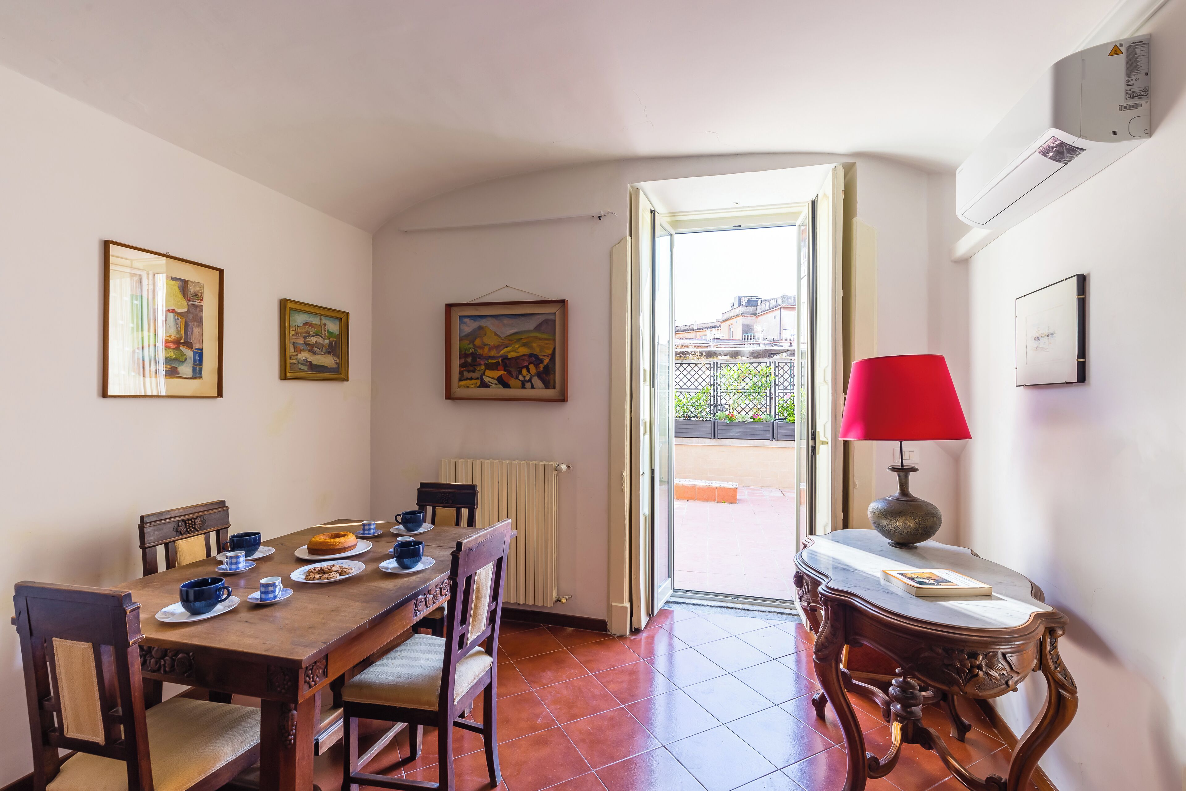 Uma sala de jantar com mesa de madeira posta para quatro pessoas, cadeiras e um abajur vermelho sobre uma mesinha lateral.