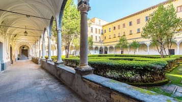 Uma galeria coberta com arcos, um pátio com sebes aparadas e um edifício com várias janelas.