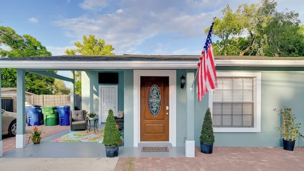 Uma casa térrea com varanda frontal, porta de madeira com painel de vidro decorativo, uma bandeira americana e uma janela com persianas.