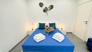 Cama bem arrumada com roupa de cama azul, duas toalhas brancas, um suporte com amenities e arte decorativa na parede.