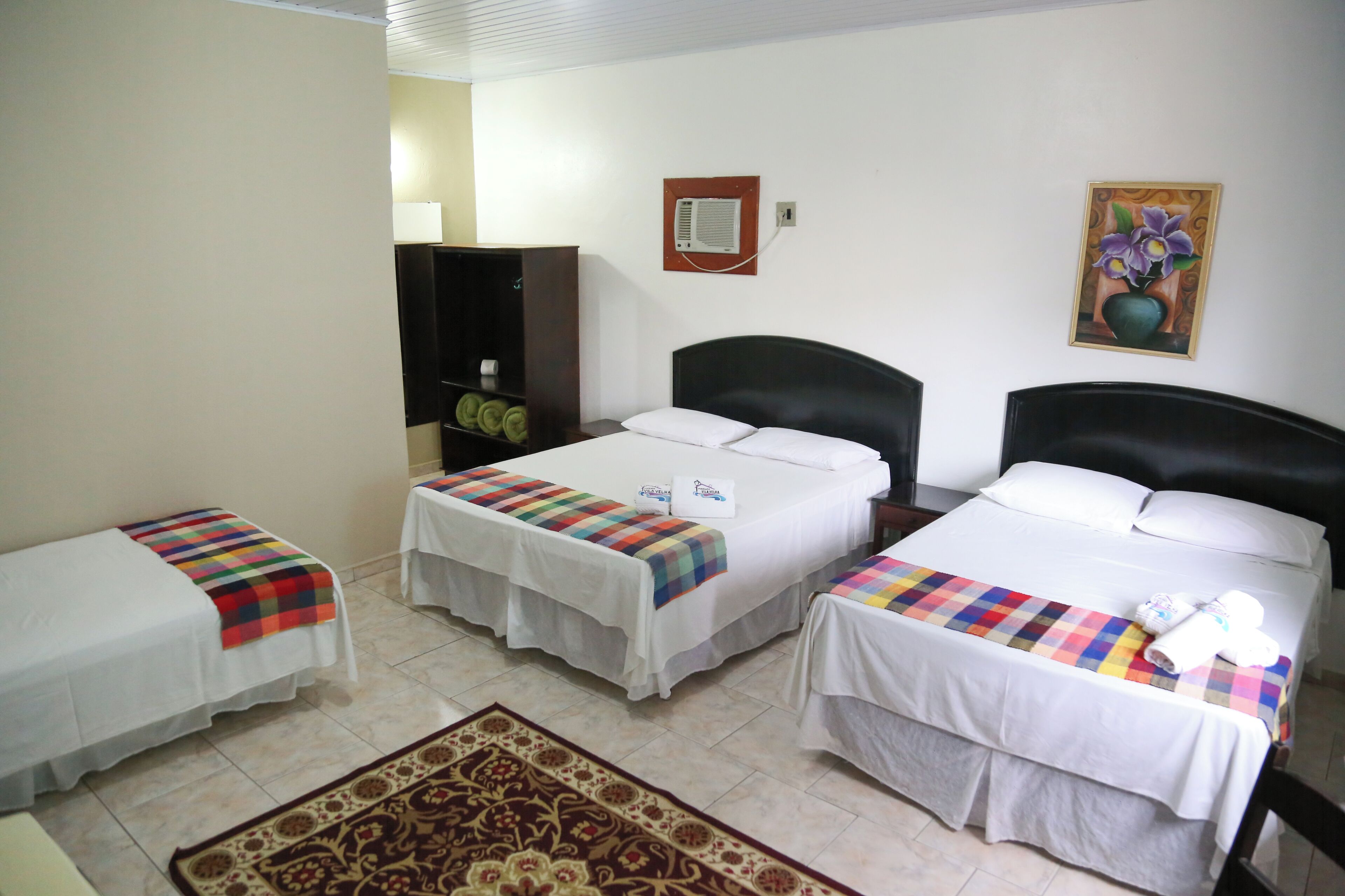 Quarto de hotel com três camas, um guarda-roupa, um quadro e uma pequena mesa com abajur.