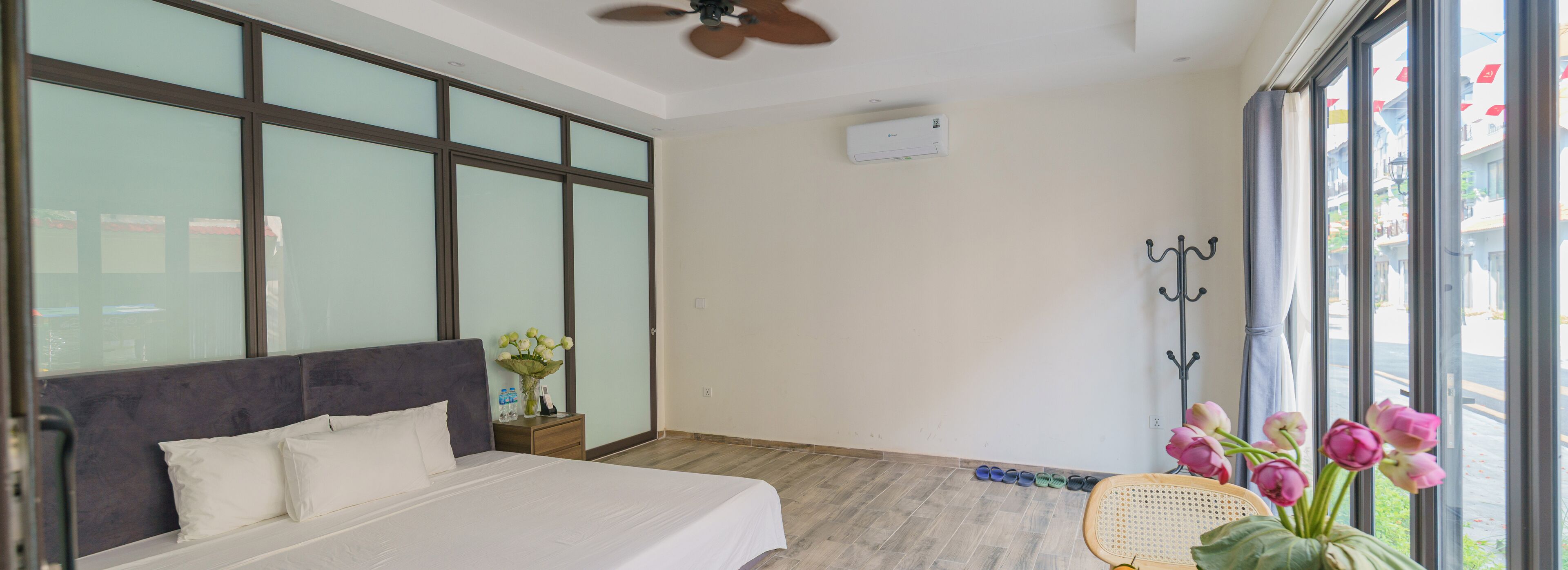 Quarto de hotel moderno com uma cama grande, ventilador de teto, janela com vista e uma mesa pequena com um vaso de flores.