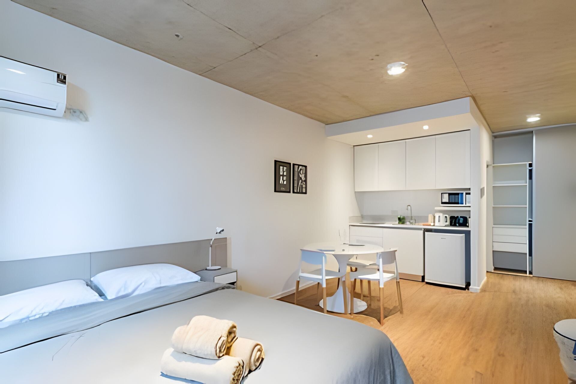 Quarto de hotel moderno com cama, mesa de cabeceira e uma pequena cozinha equipada.