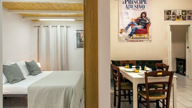 Quarto com cama, área de refeições com mesa e cadeiras, e um cartaz na parede.