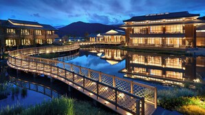 Um hotel de estilo tradicional com vários edifícios interligados por uma ponte sobre uma superfície aquática calma, rodeado por paisagens naturais.