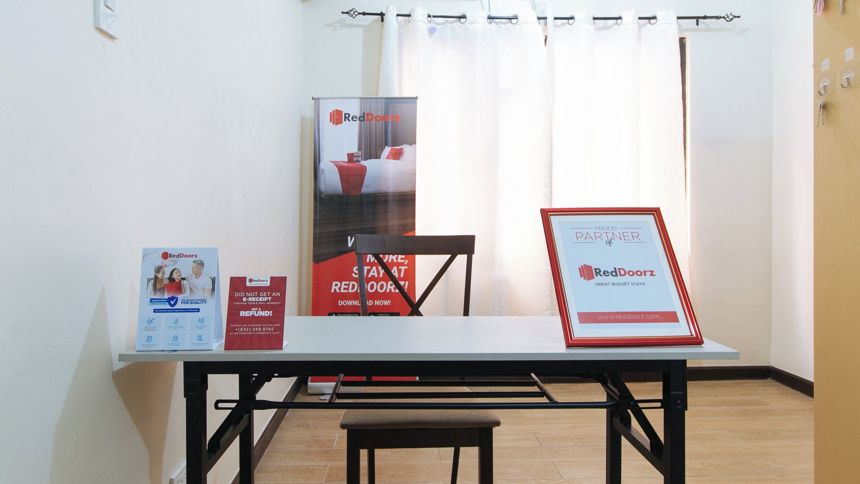 Quarto com uma mesa exibindo material promocional da RedDoorz, uma cadeira e uma cortina.