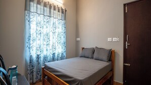 Quarto com cama de madeira, roupa de cama cinza, janela com cortinas florais, mesa de cabeceira com garrafa de água azul e porta de madeira escura.