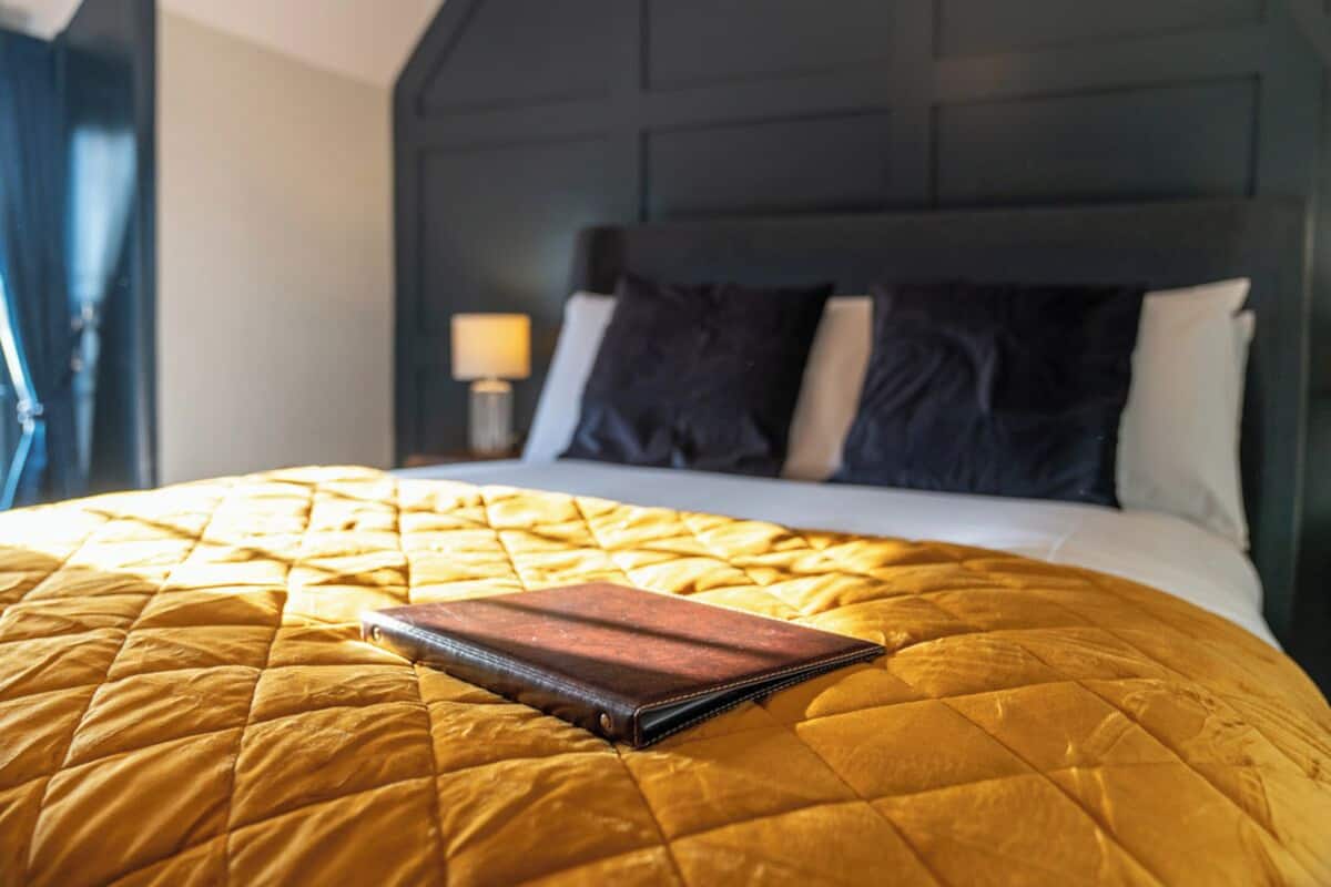 Cama bem arrumada com colcha amarela, um objeto de couro marrom sobre a cama e uma cabeceira escura.