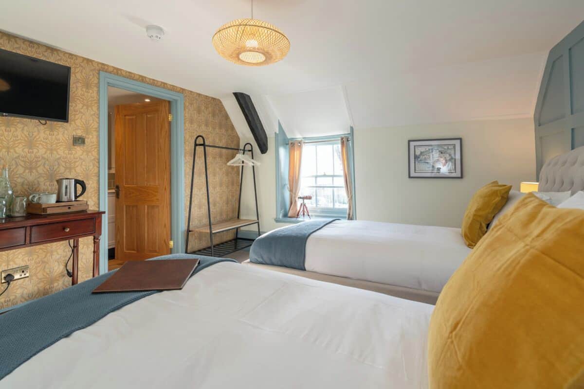 Quarto de hotel com duas camas, uma escrivaninha com chaleira, televisão de tela plana e uma janela com cortinas.