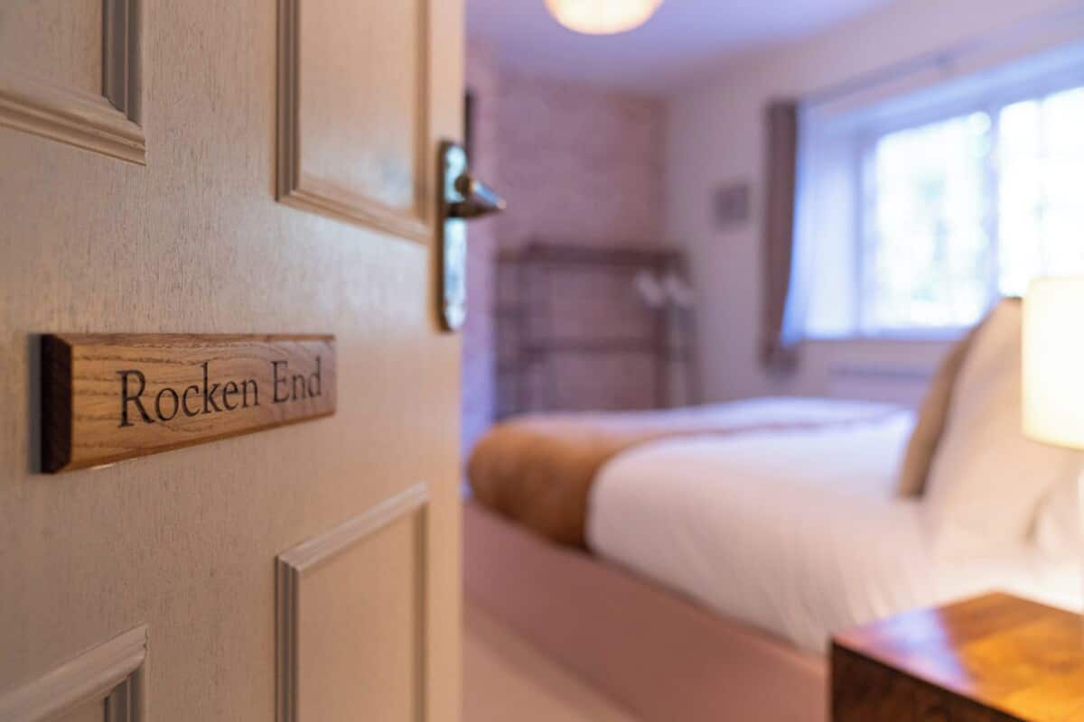 Quarto de hotel com cama, mesa de cabeceira, janela e porta com a placa 'Rocken End'.