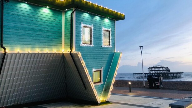 Um edifício cor de verde-água com telhado inclinado, duas janelas com molduras iluminadas e um caminho pavimentado que leva a um píer.