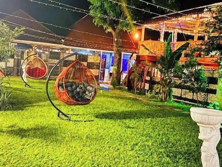 Uma área externa bem iluminada com gramado, cadeiras suspensas e um edifício com telhado de palha.