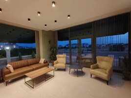 Uma sala de estar moderna com um sofá, duas poltronas e uma mesa de centro. Grandes janelas oferecem vista para a cidade ao entardecer.