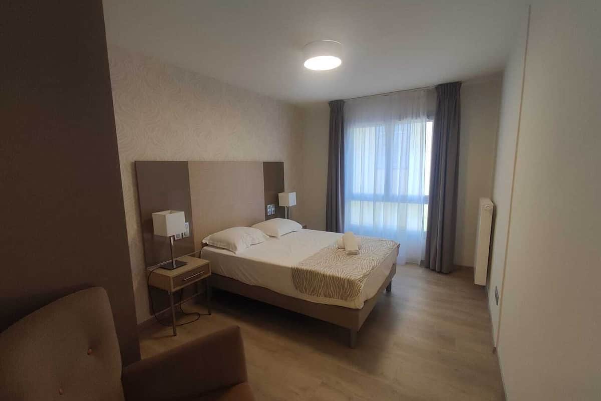 Quarto de hotel com uma cama grande, mesas de cabeceira, uma cadeira e uma janela com cortinas.