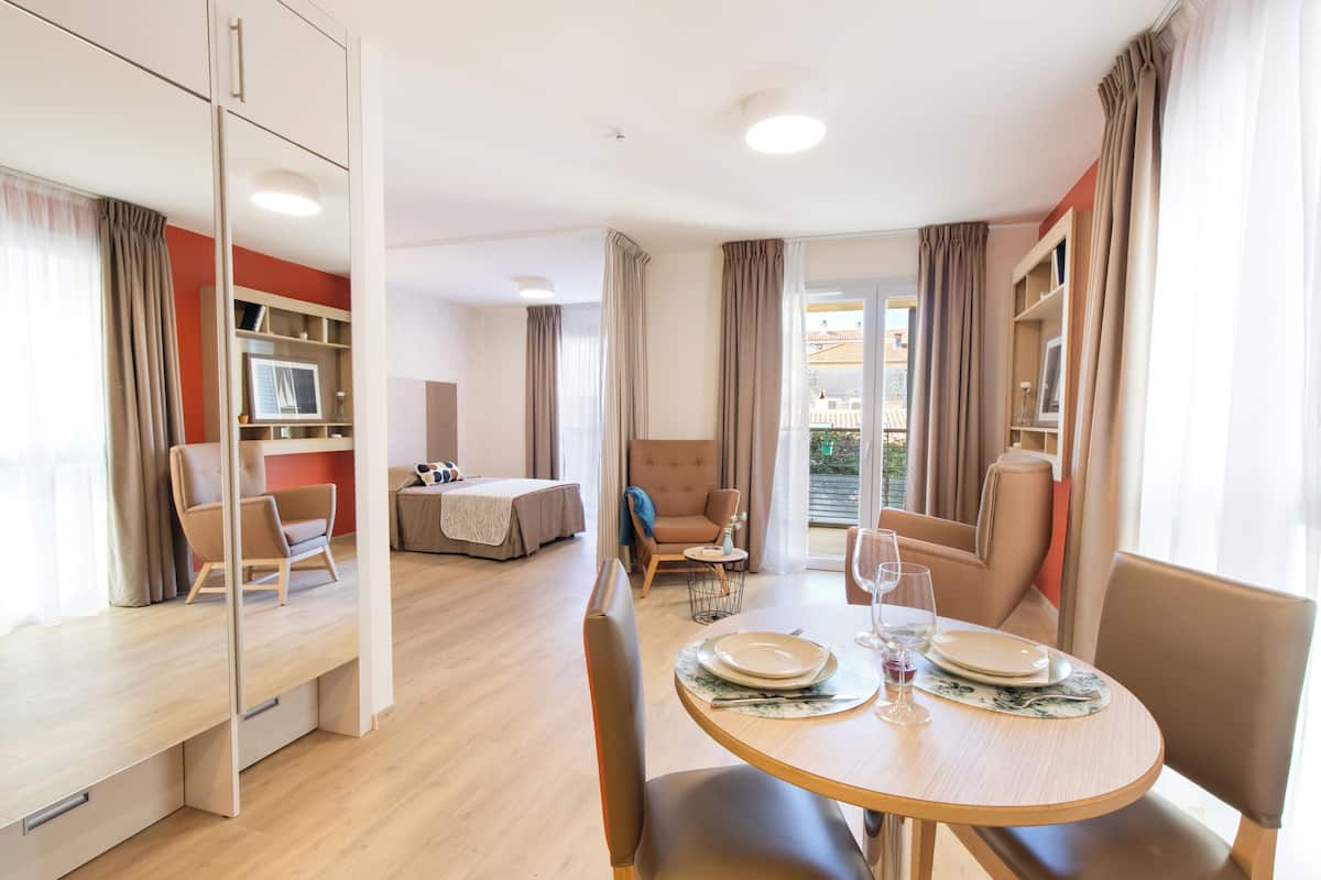 Quarto de hotel moderno com área de jantar, sofá, poltrona e televisão.