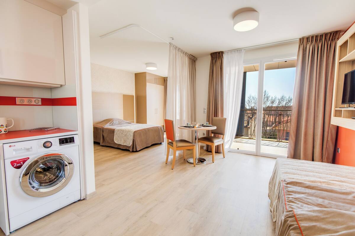 Quarto de hotel com máquina de lavar, uma cama, mesa de jantar com cadeiras e varanda com cortinas.