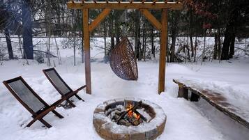 Uma área externa coberta de neve, com um braseiro, cadeiras de madeira e uma estrutura de pérgola.