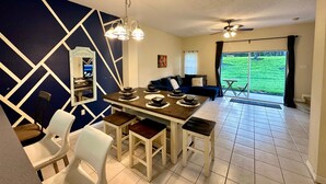 Uma sala de jantar com mesa e cadeiras, um ventilador de teto e vista para um campo verde através de uma porta de correr.