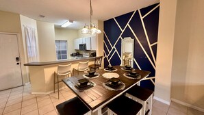 Uma cozinha moderna com armários brancos, micro-ondas e fogão. Há uma área de jantar com mesa de madeira, quatro cadeiras e um espelho decorativo na parede.
