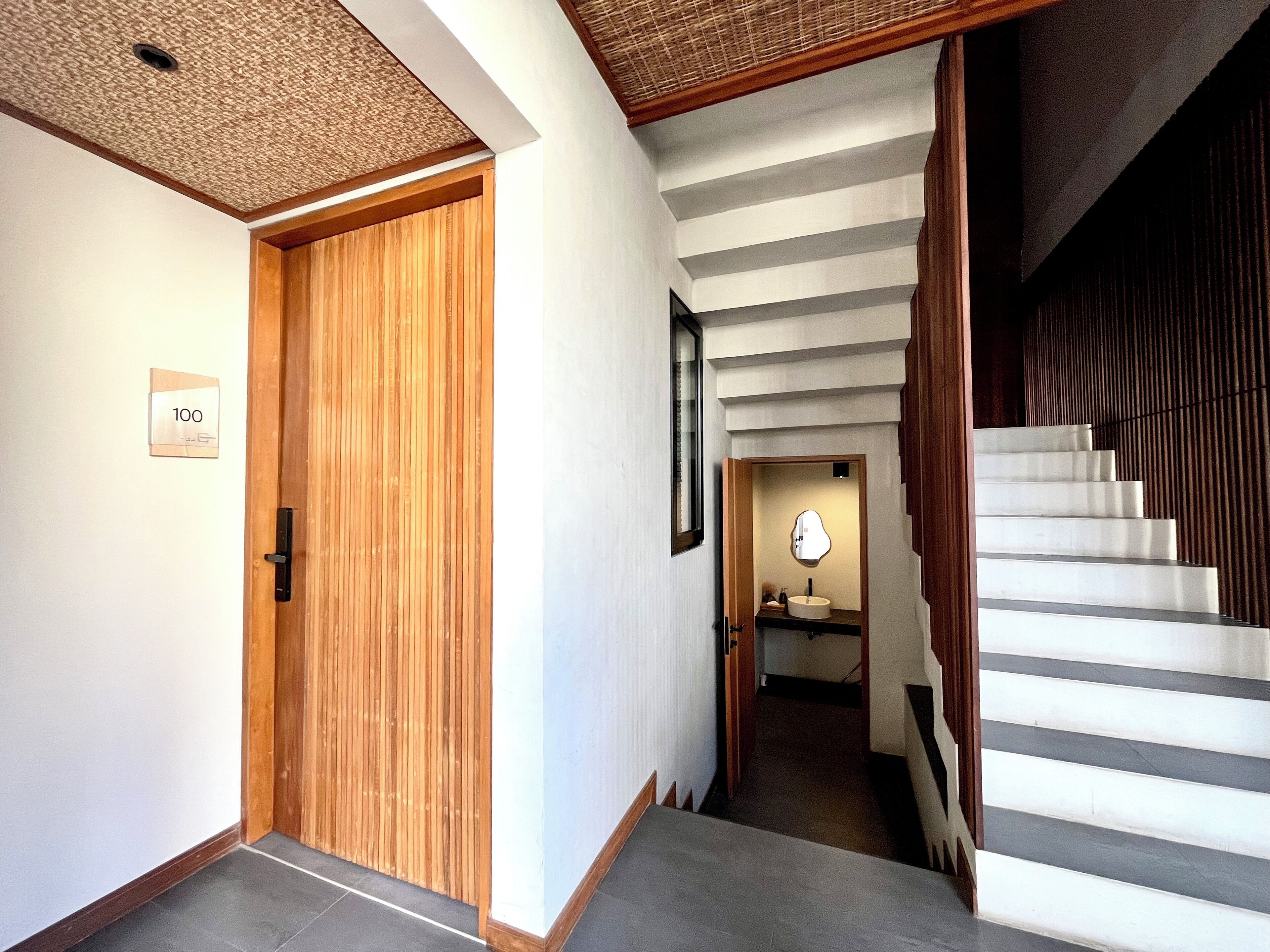 Um corredor com porta de madeira marcada '100', escada com corrimão escuro e um pequeno banheiro com espelho e pia.