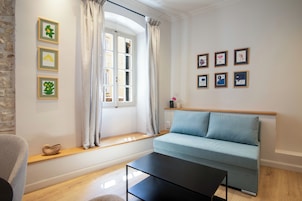Uma sala de estar moderna com um sofá azul claro, uma mesa de centro preta e uma janela com cortinas brancas. Há quadros nas paredes e uma prateleira de madeira com objetos decorativos.