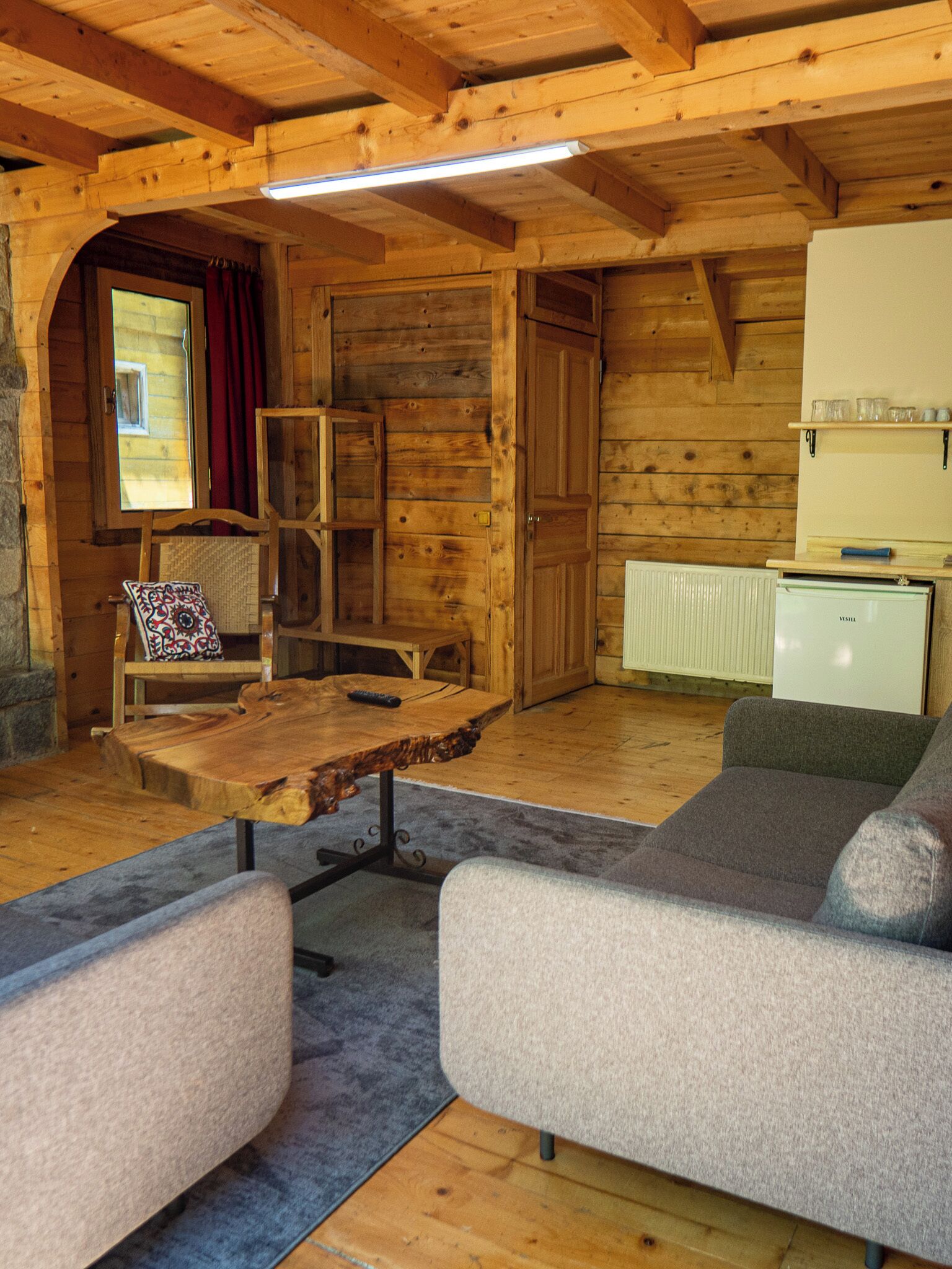 Interior acolhedor de cabana com mesa de jantar de madeira, dois sofás cinza, pequena cozinha com geladeira e janela com cortinas vermelhas.