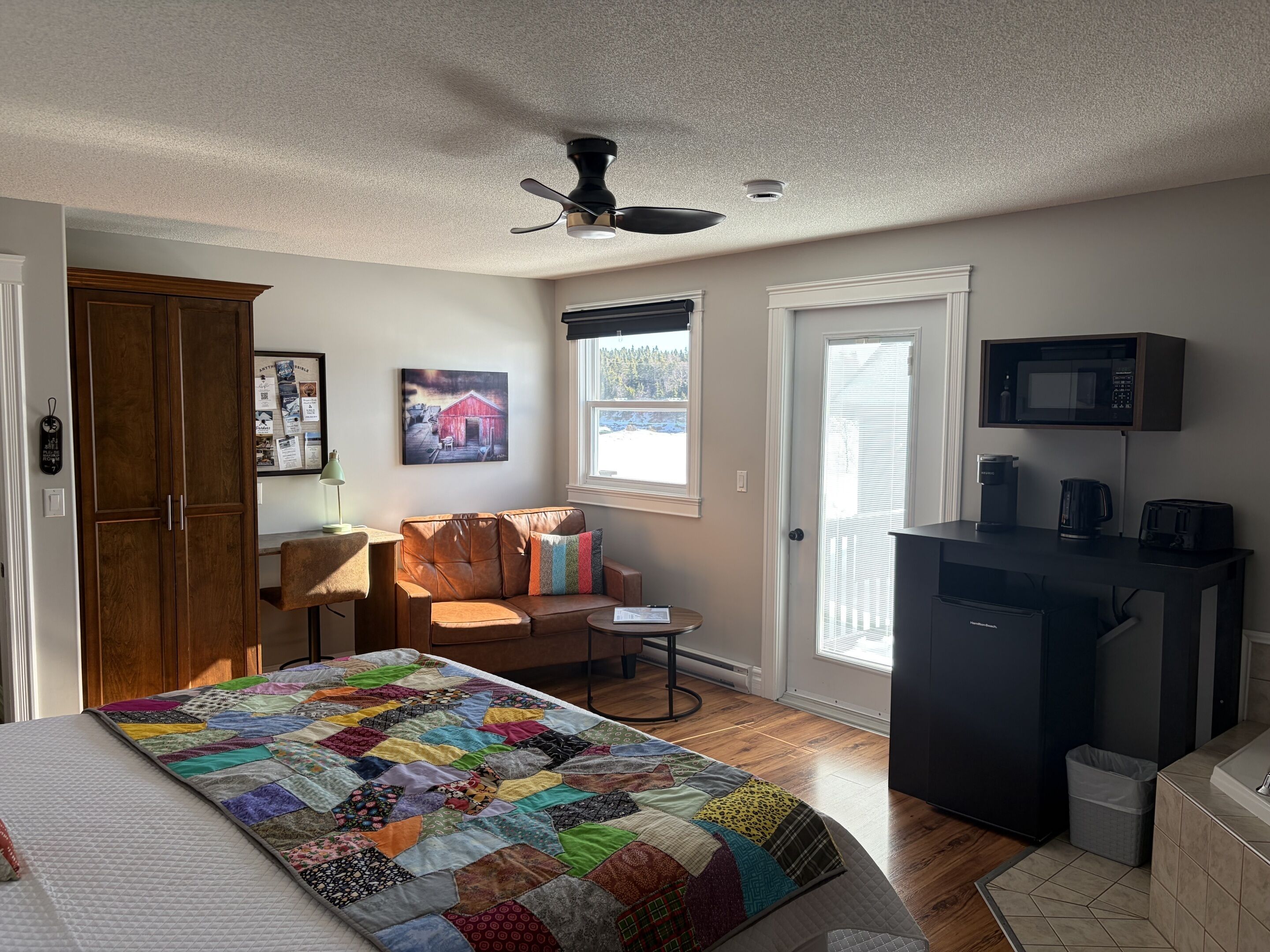 Quarto com cama, escrivaninha, cadeira, ventilador de teto, janela e kitchenette.