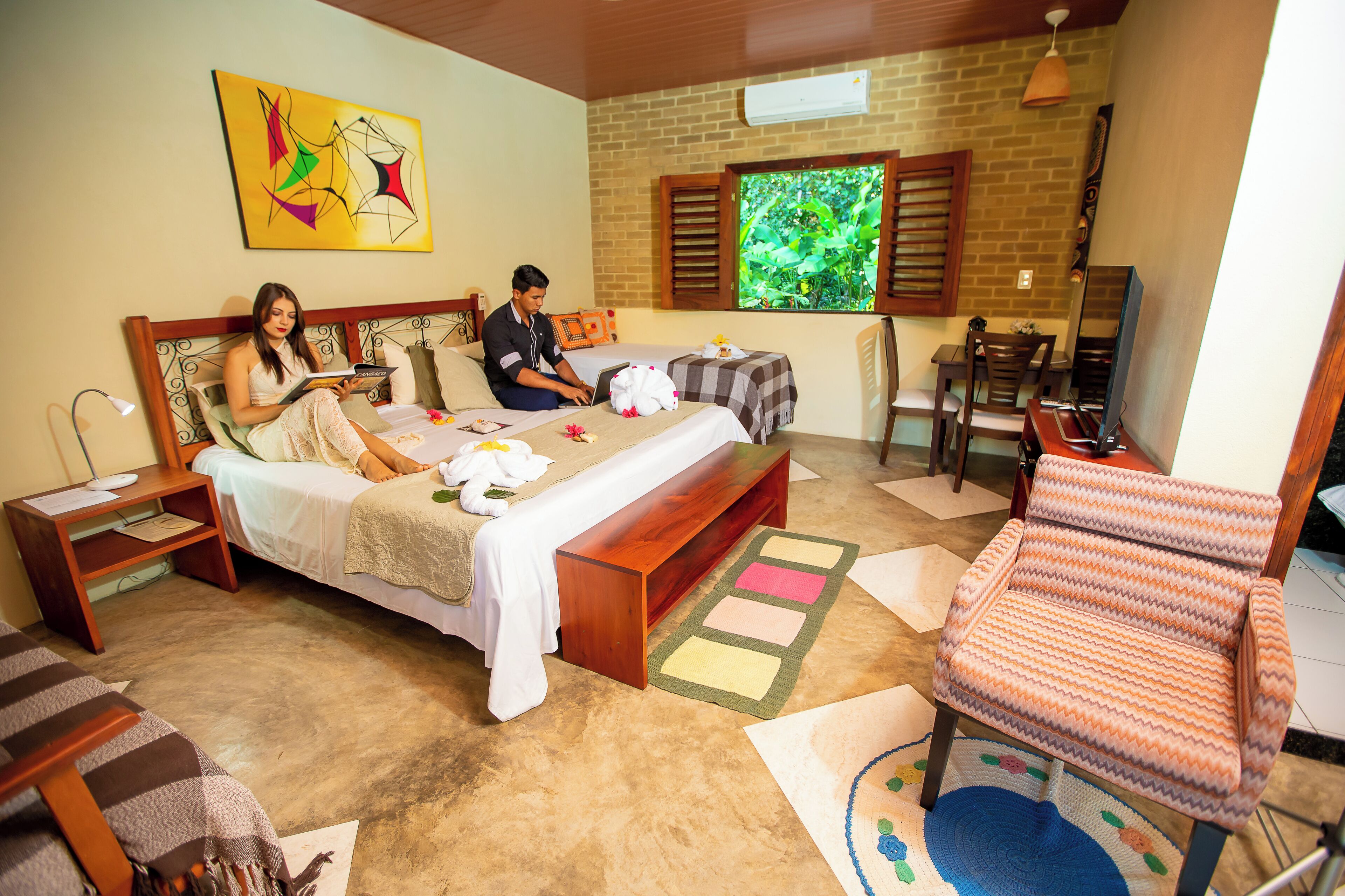 Quarto de hotel com uma cama grande, uma cama menor, uma escrivaninha, uma cadeira e uma janela com vista para o verde.
