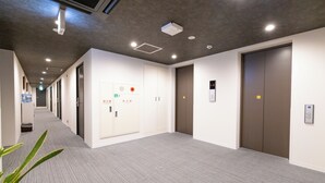 Um corredor com carpete cinza, paredes brancas e teto com iluminação embutida. Há portas à direita, uma delas com botão de elevador.