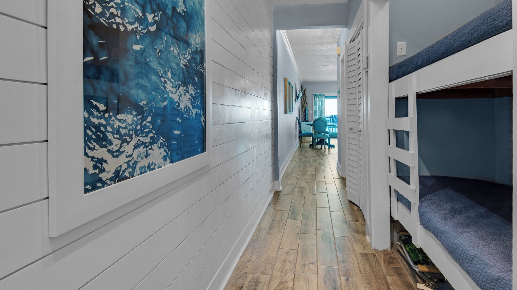 Um corredor com beliches, uma grande pintura abstrata na parede esquerda e vista para um quarto com uma cadeira azul e uma mesa.