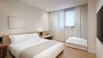 Quarto de hotel moderno com cama grande, mesa de cabeceira, televisão de tela plana e janela com cortinas leves.