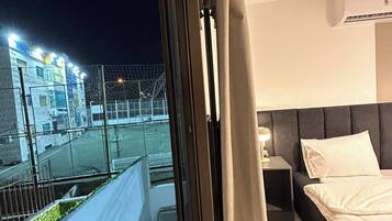 Quarto com cama, mesa de cabeceira, ar condicionado e vista para um campo esportivo através de uma janela.