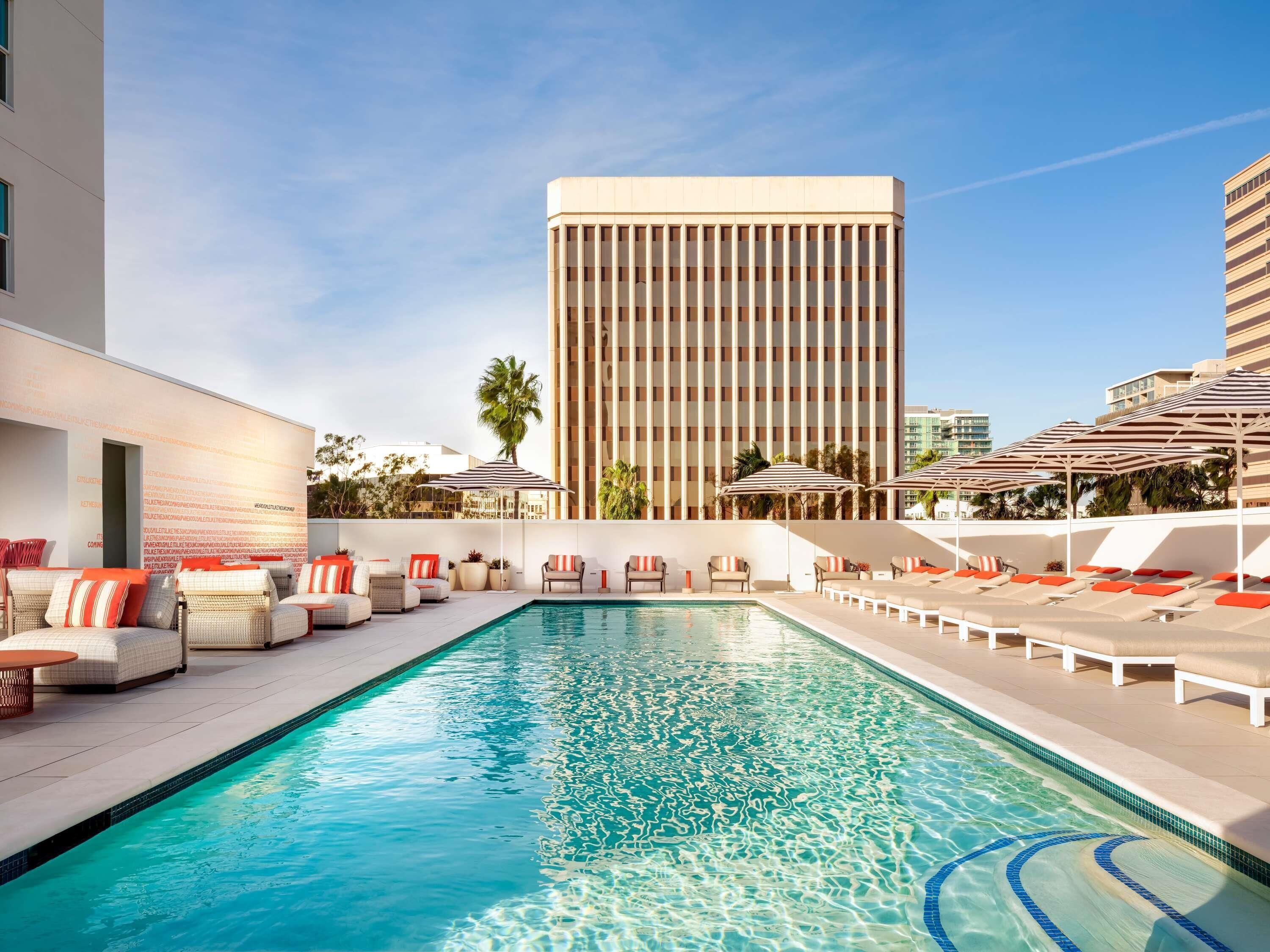 Piscina no terraço com espreguiçadeiras e guarda-sóis, rodeada por um edifício moderno com fachada vertical.