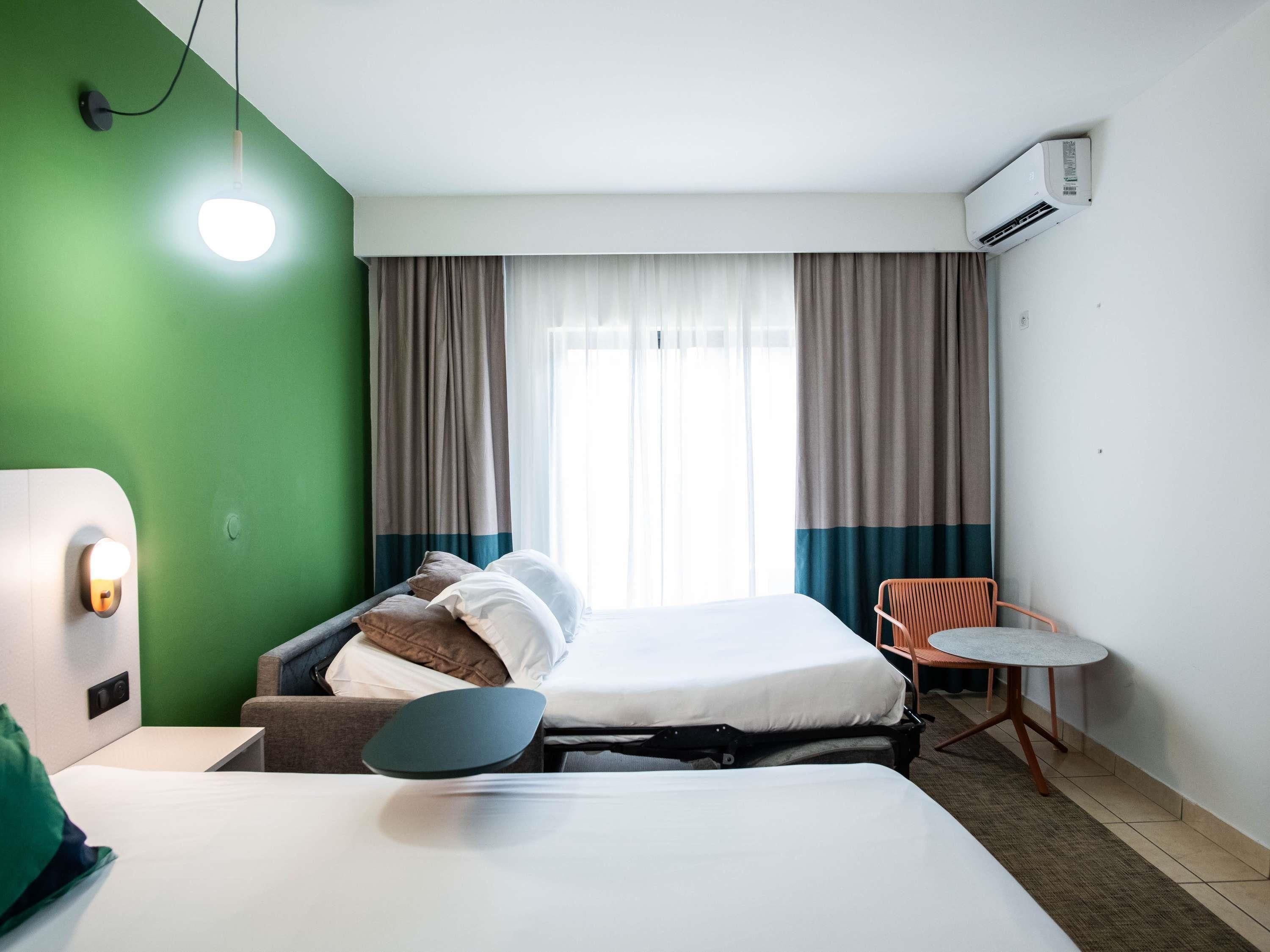 Quarto de hotel com cama, uma cadeira, uma mesinha e janela com cortinas.