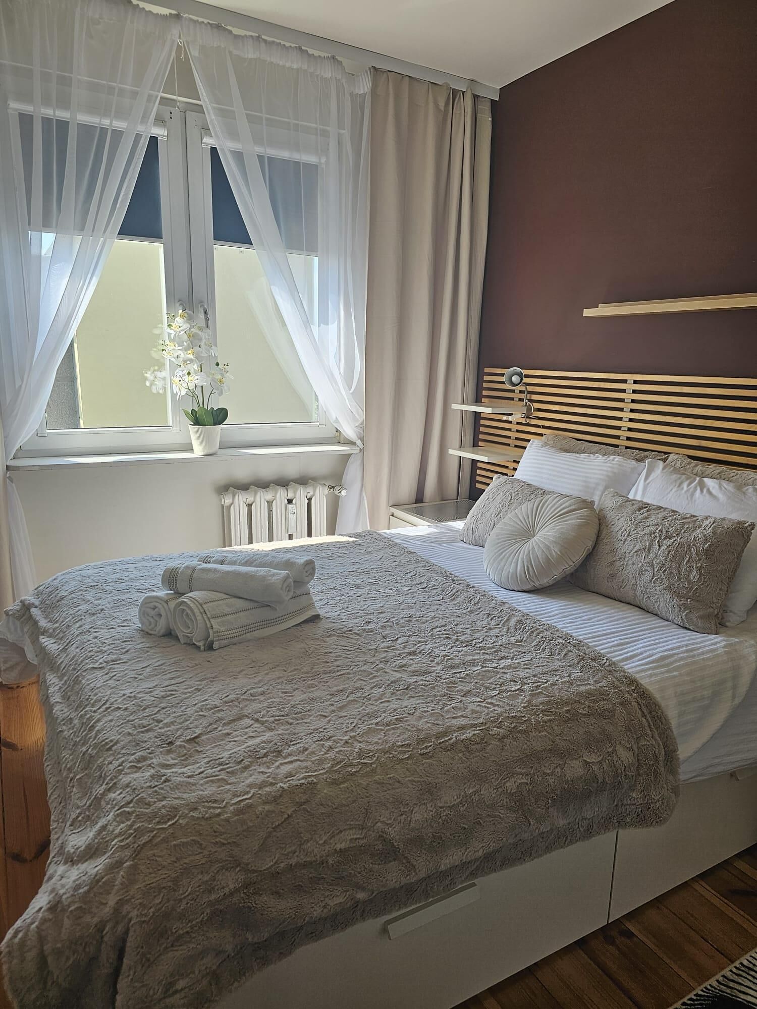 Uma cama bem arrumada com manta e almofadas texturizadas, uma mesinha de cabeceira com abajur, e uma janela com cortinas finas e planta em vaso.
