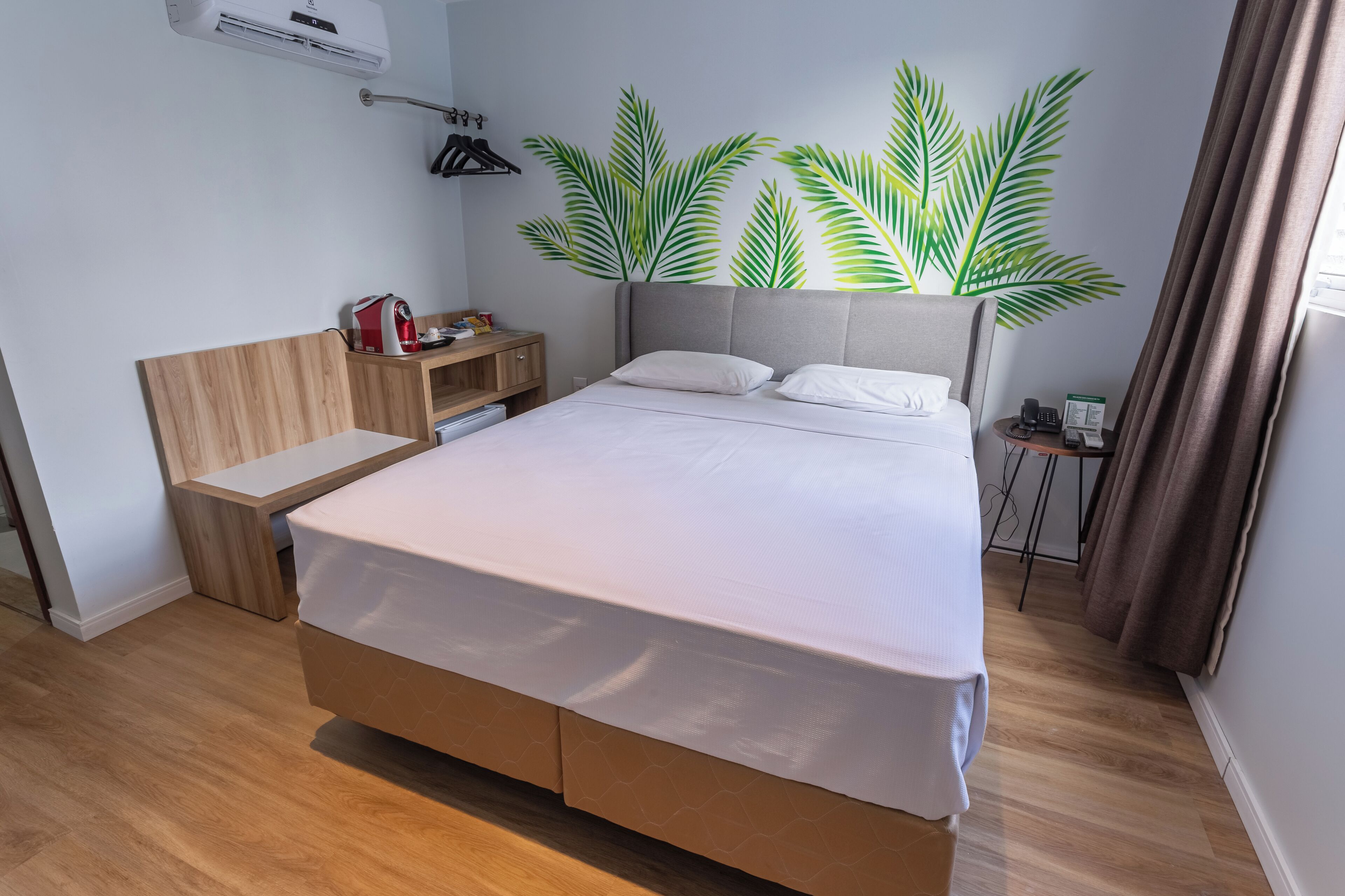Quarto de hotel com cama, mesa de cabeceira e uma pequena escrivaninha com cadeira. Há um ar condicionado fixado na parede e uma decoração de folhas verdes.