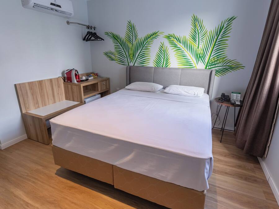 Quarto de hotel com cama, mesa de cabeceira e uma pequena escrivaninha com cadeira. Há um ar condicionado fixado na parede e uma decoração de folhas verdes.