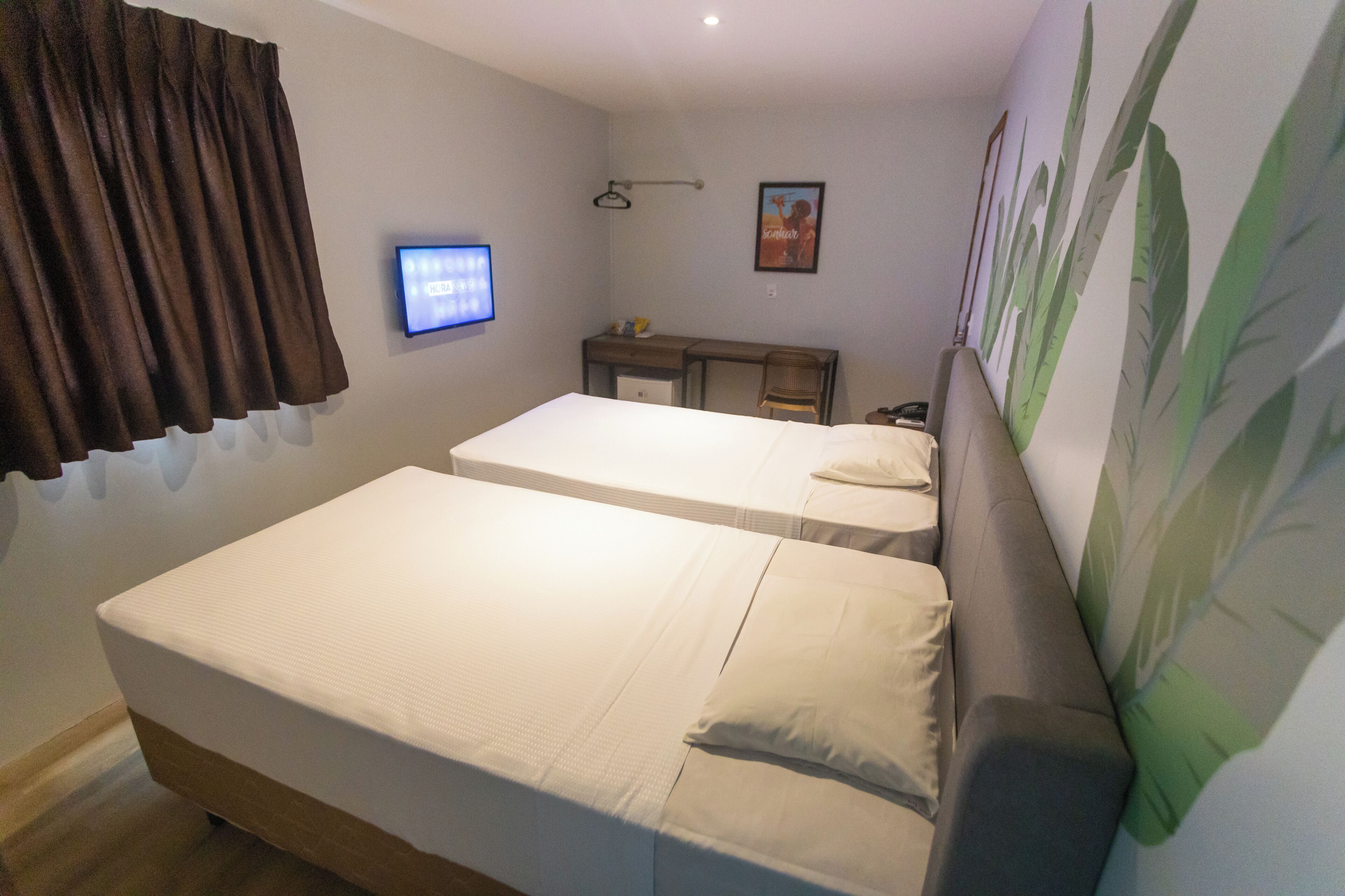 Quarto com duas camas, uma escrivaninha, uma cadeira, uma televisão e uma parede com padrão de folhas.