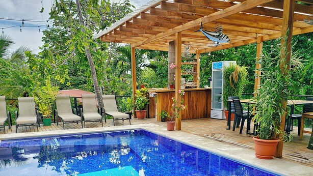 Piscina com pergolado de madeira, cercada por plantas em vasos e mobiliário para área externa.