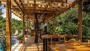 Um deque de madeira ao ar livre com mesas e cadeiras, cercado por vegetação exuberante e uma piscina.