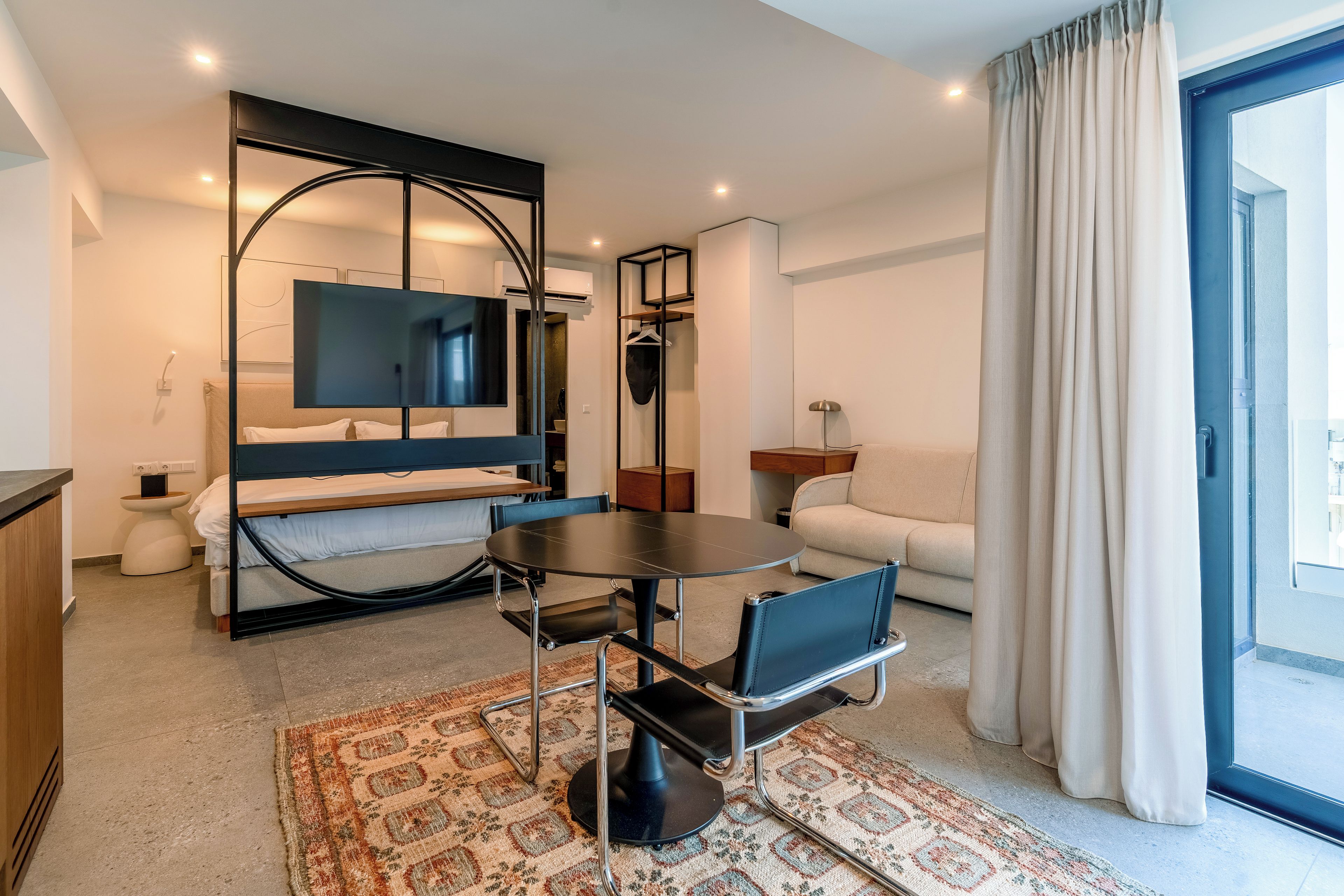 Quarto de hotel moderno com sofá, mesa de jantar com cadeiras, televisão e uma grande janela com cortinas.