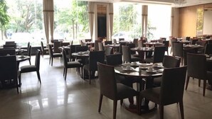 Um restaurante com mesas e cadeiras redondas, janelas amplas com cortinas e um candelabro.